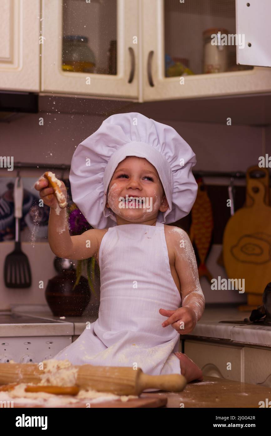 A kid in a cook's costume sits in the kitchen on a table and smiles ...