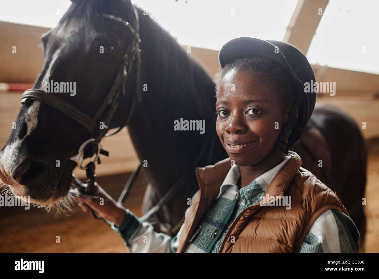 Portrait of young black woman posing with horse in indoor riding arena ...
