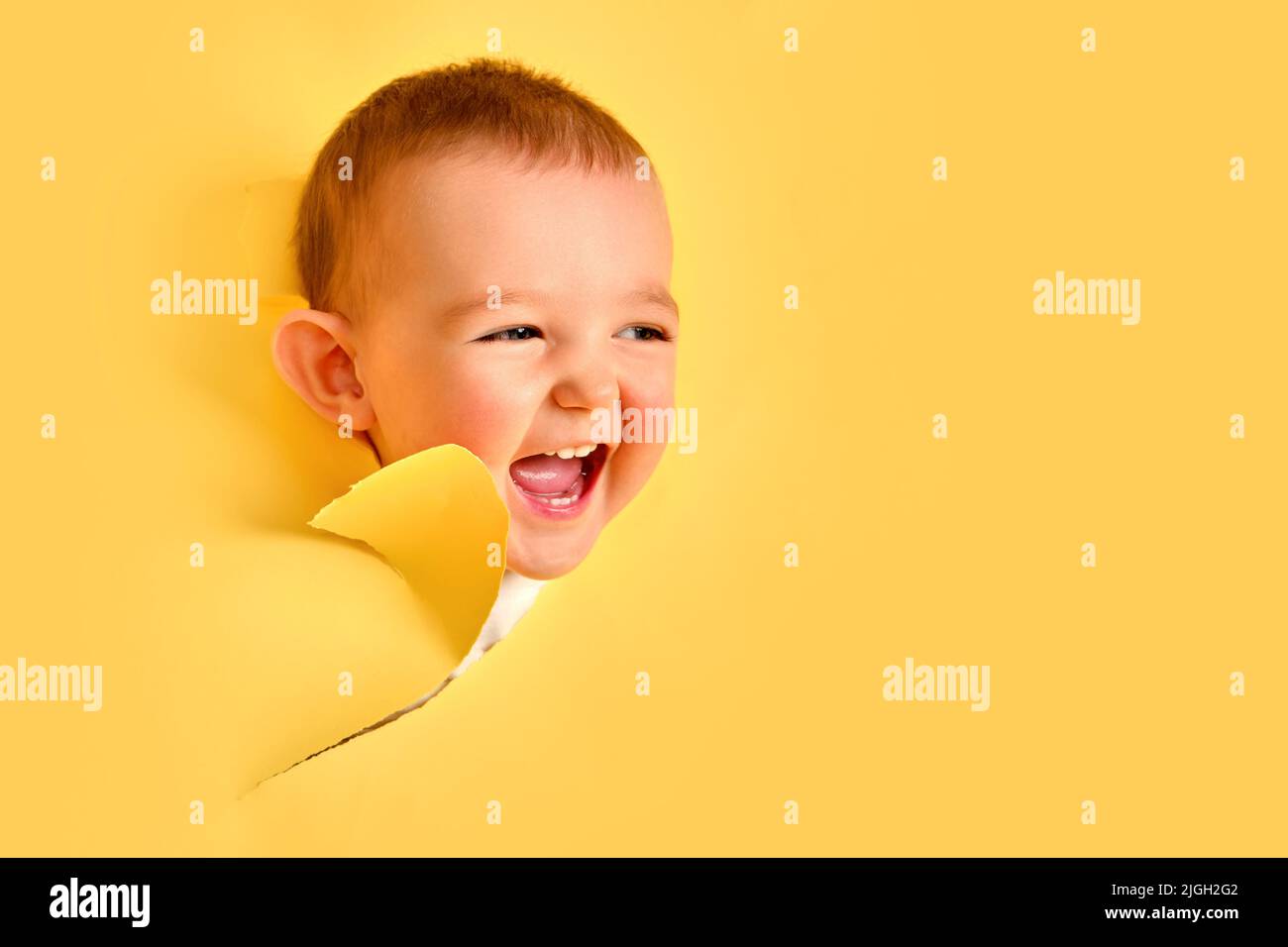 A happy child looks out of a hole in the studio yellow background ...