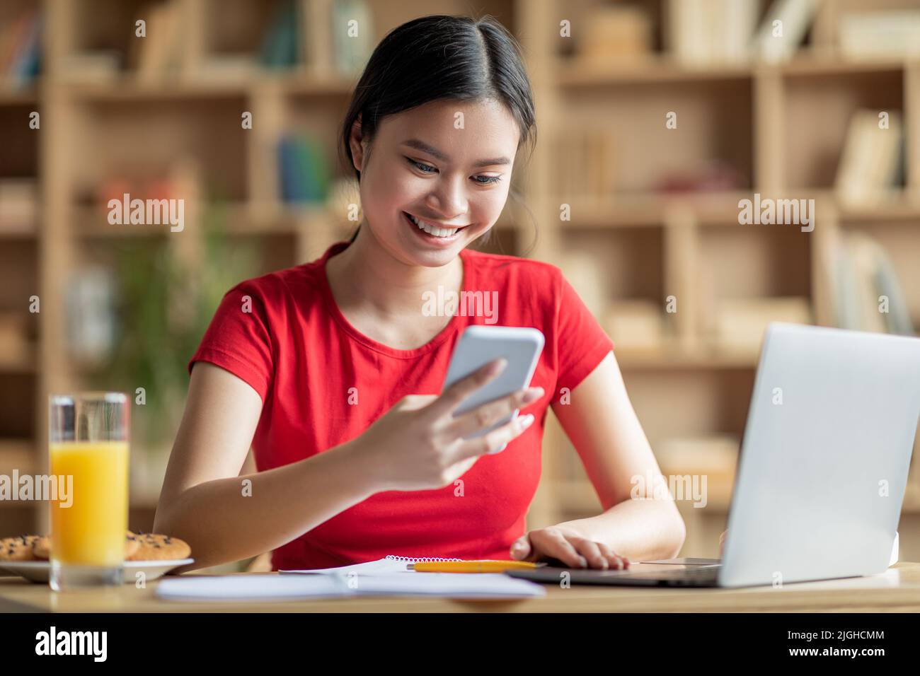 Cheerful adolescent chinese lady student typing on smartphone at table ...