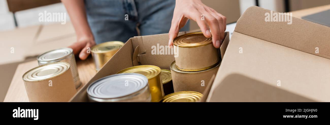 cropped view of african american volunteer packing canned food in box ...