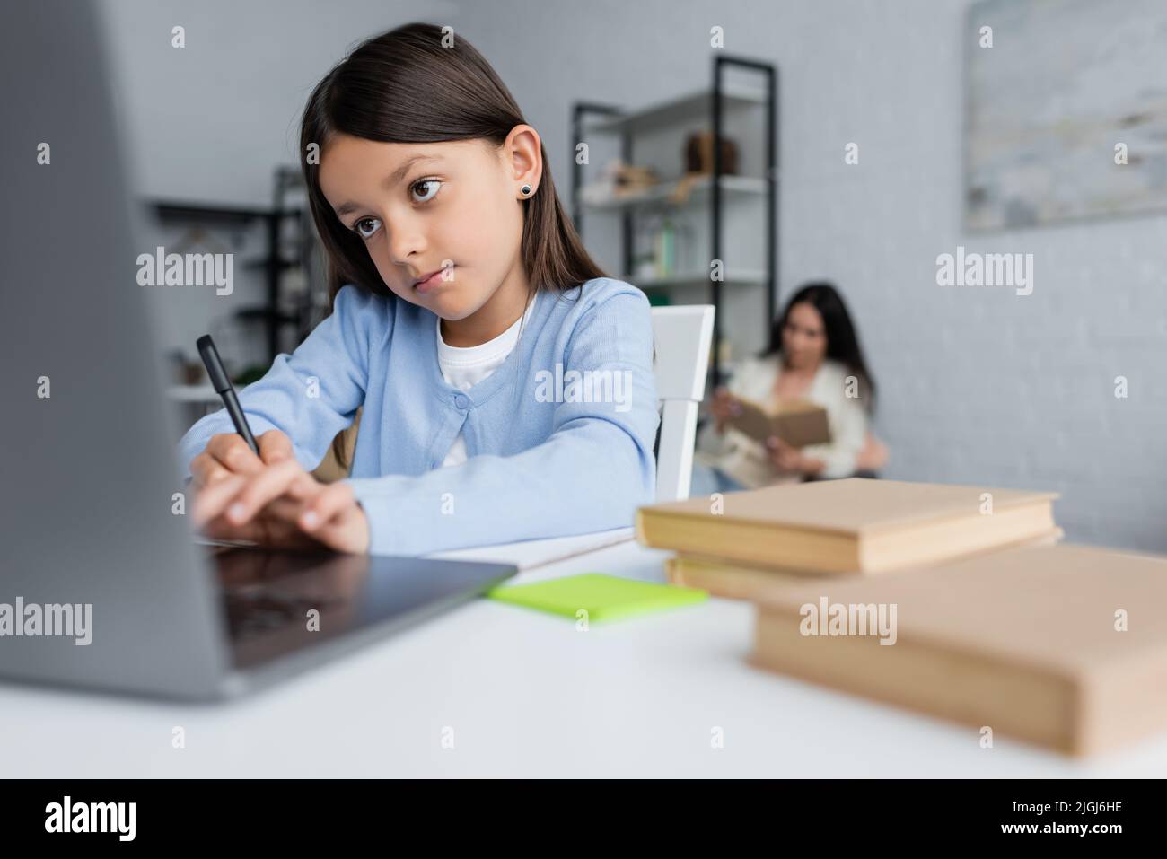 girl writing near blurred laptop while doing homework near nanny on ...