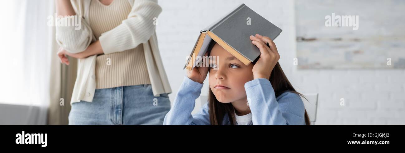 bored and tired girl covering head with textbook near babysitter on ...