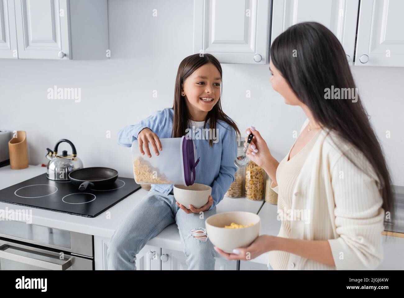 happy girl sitting on kitchen counter and pouring corn flakes into bowl ...