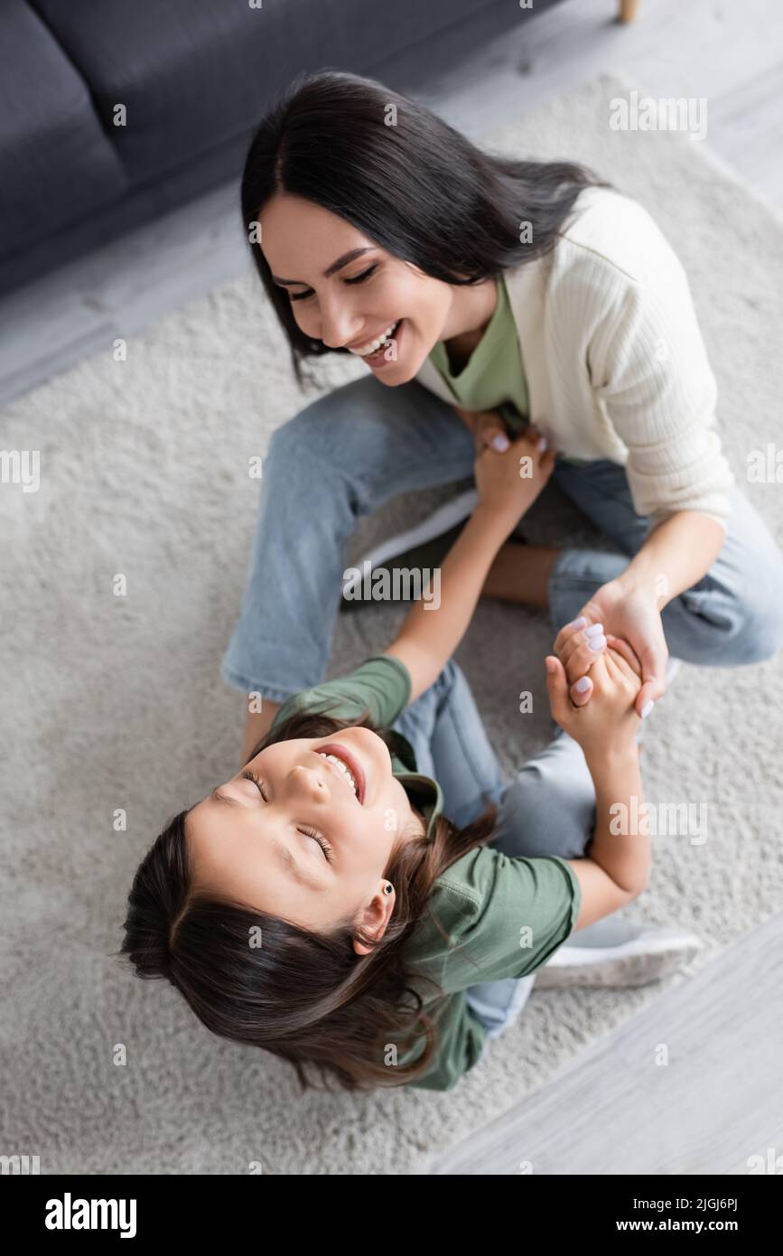 top view of cheerful babysitter and girl having fun on carpet at home ...