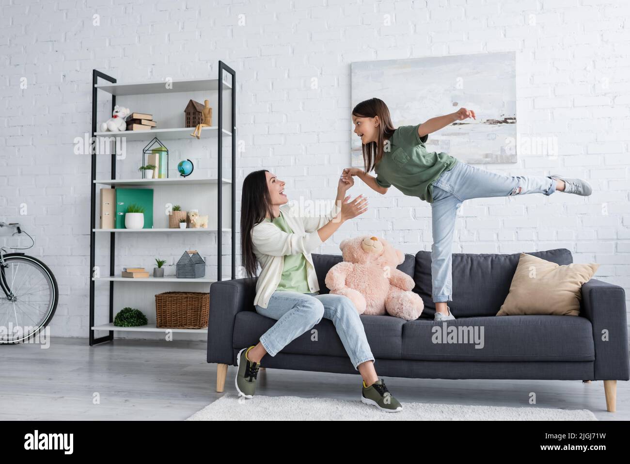 happy babysitter supporting excited kid standing on one leg on couch in ...