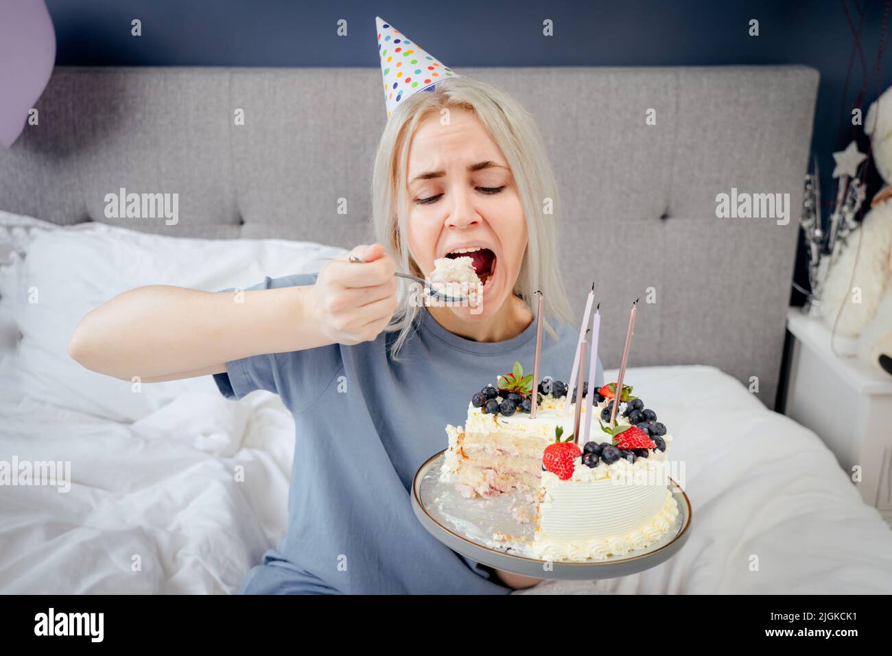 Sad, upset woman in pajama and party cap eating by spoon her birthday ...
