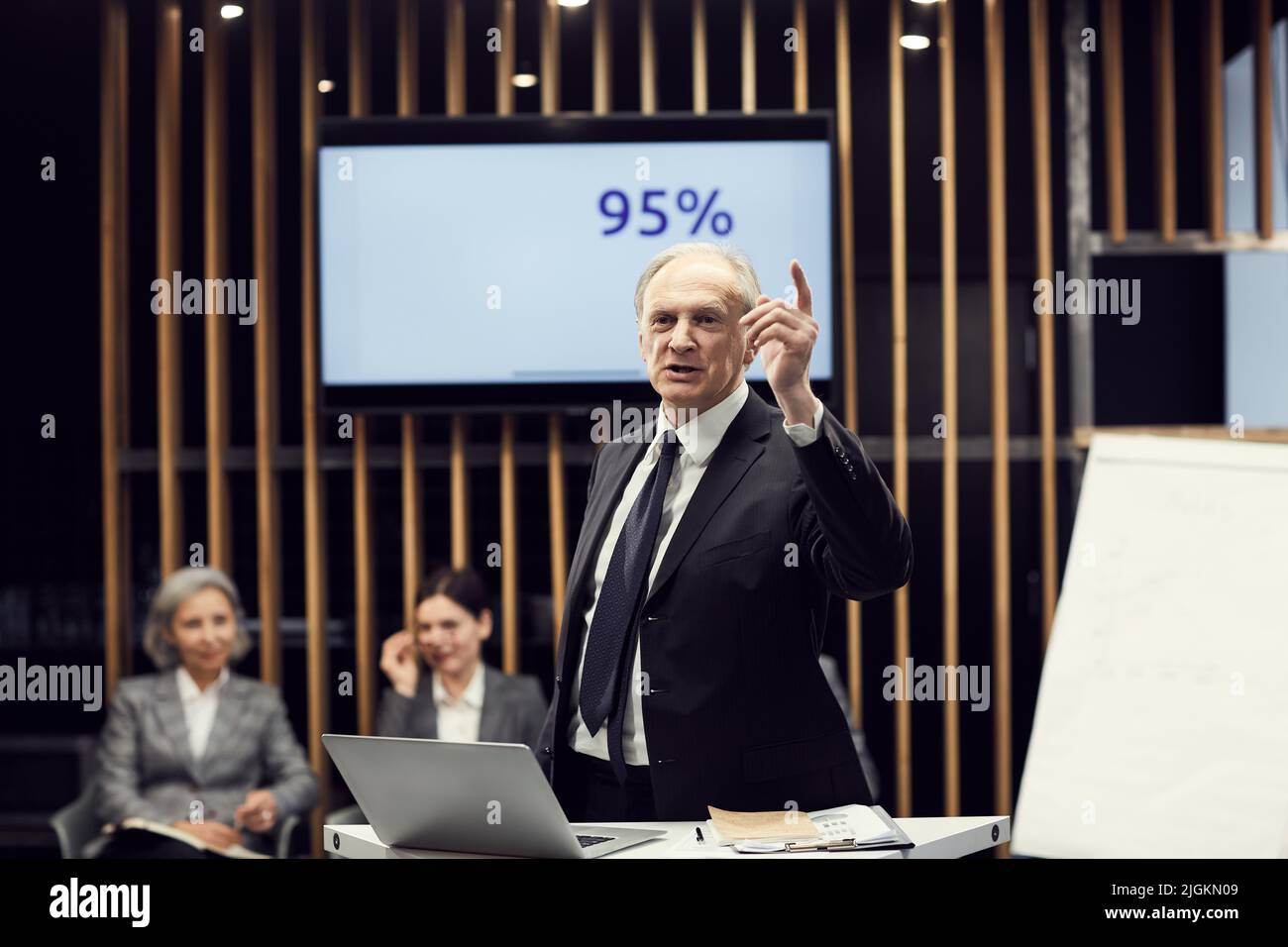 Confident senior speaker in formal suit standing at speech table with ...