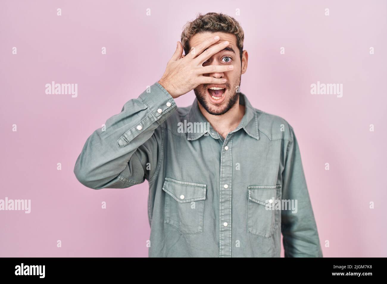 Hispanic man with beard standing over pink background peeking in shock ...