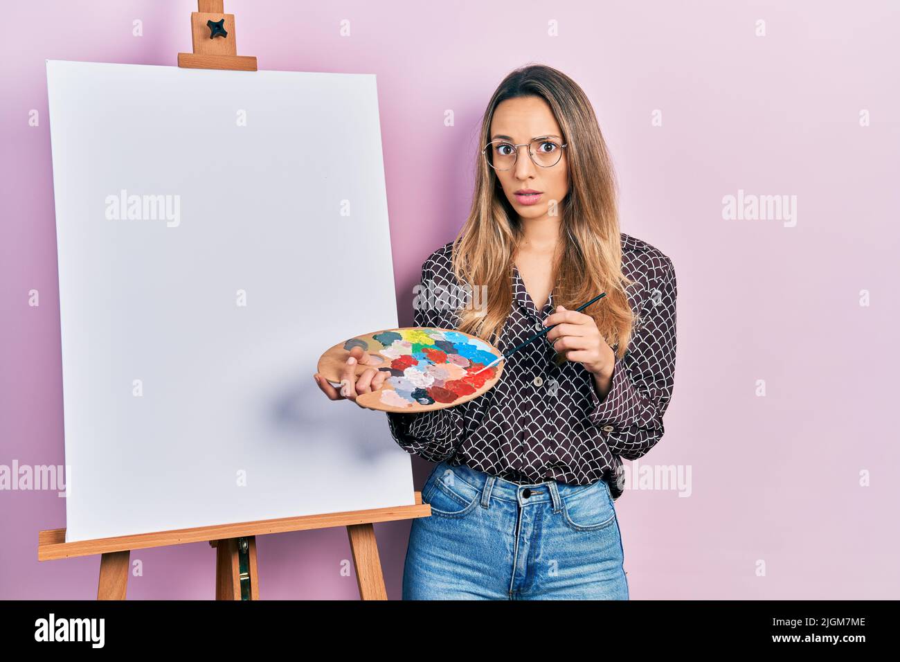 Beautiful hispanic woman standing drawing with palette by painter easel ...