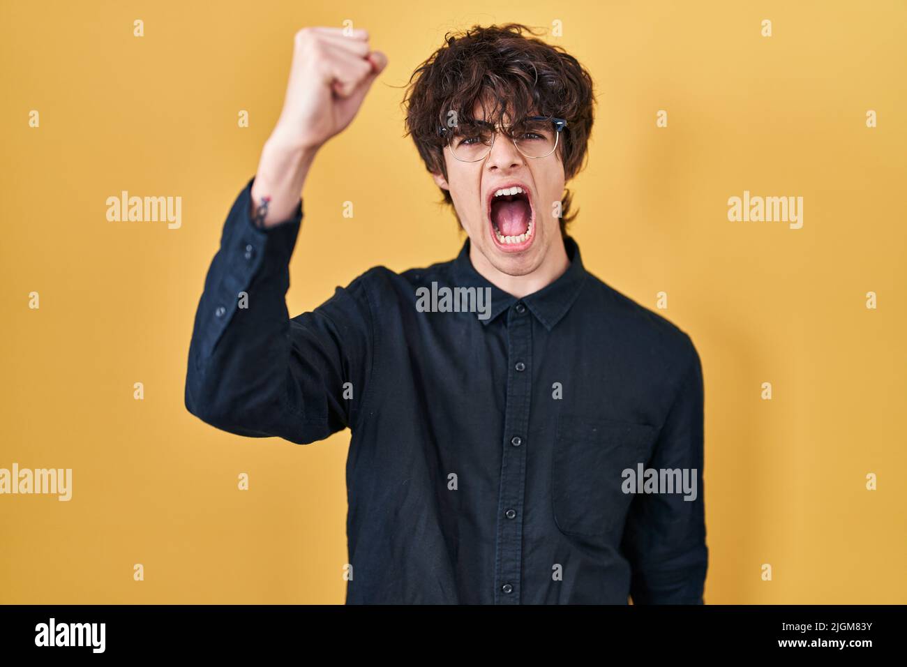 Young man wearing glasses over yellow background angry and mad raising ...