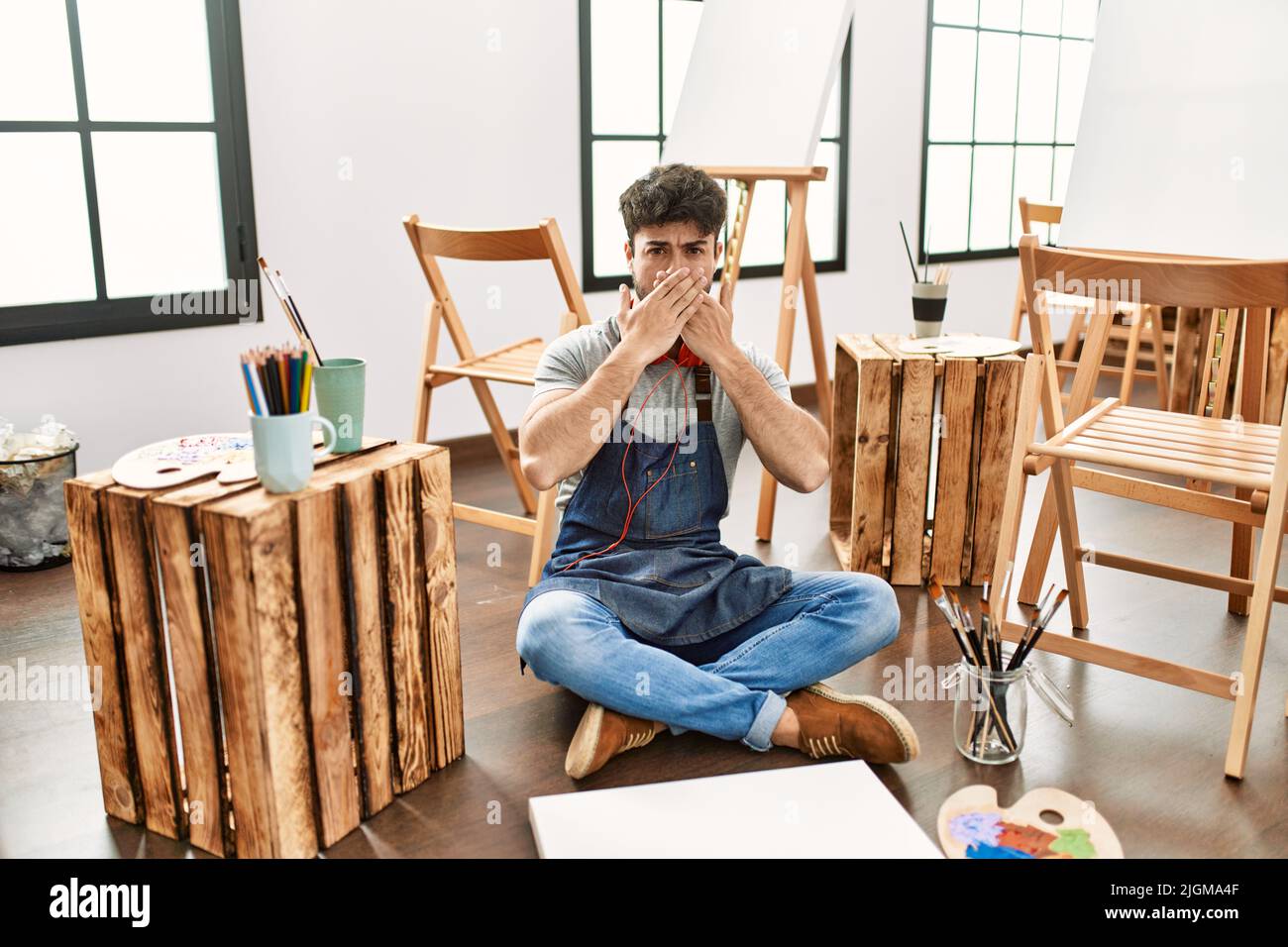 Young hispanic man sitting at art studio shocked covering mouth with ...