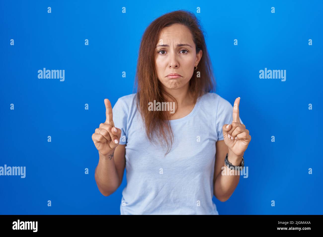 Brunette woman standing over blue background pointing up looking sad ...