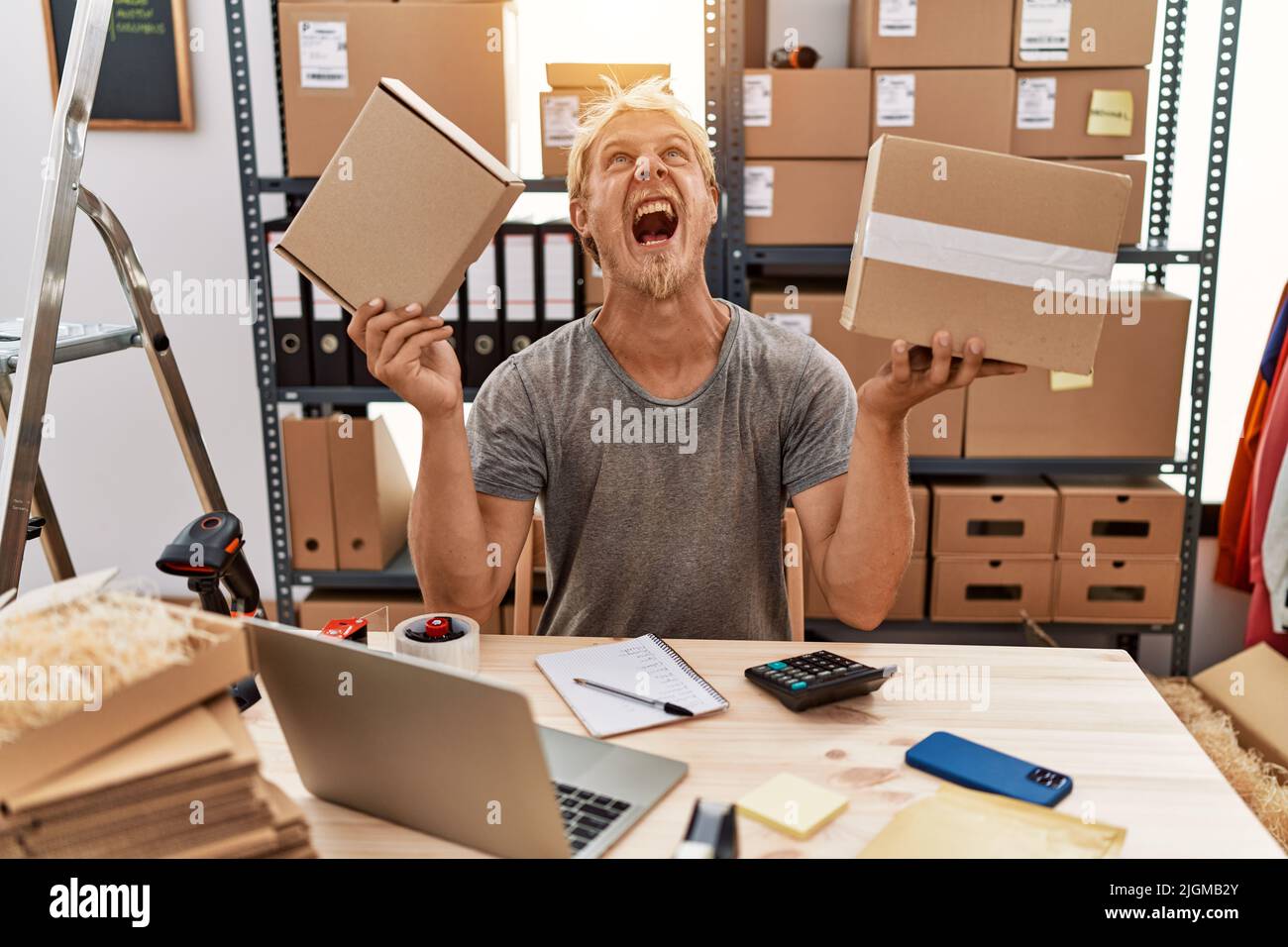 Young blond man holding packages working at online shop angry and mad ...