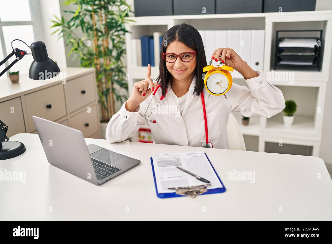 Young hispanic doctor woman holding alarm clock at the clinic surprised ...