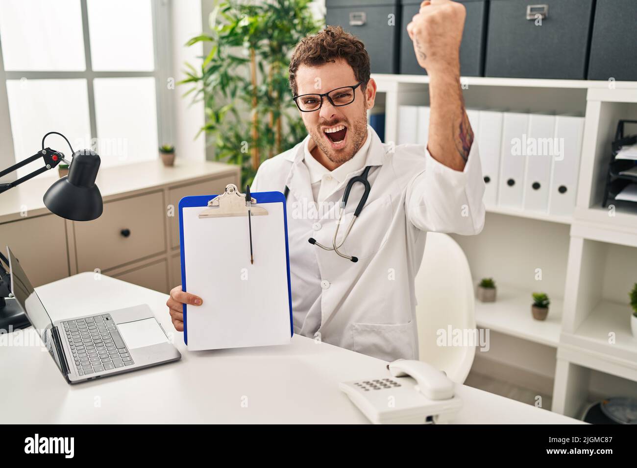 Young hispanic man wearing doctor stethoscope holding clipboard annoyed ...