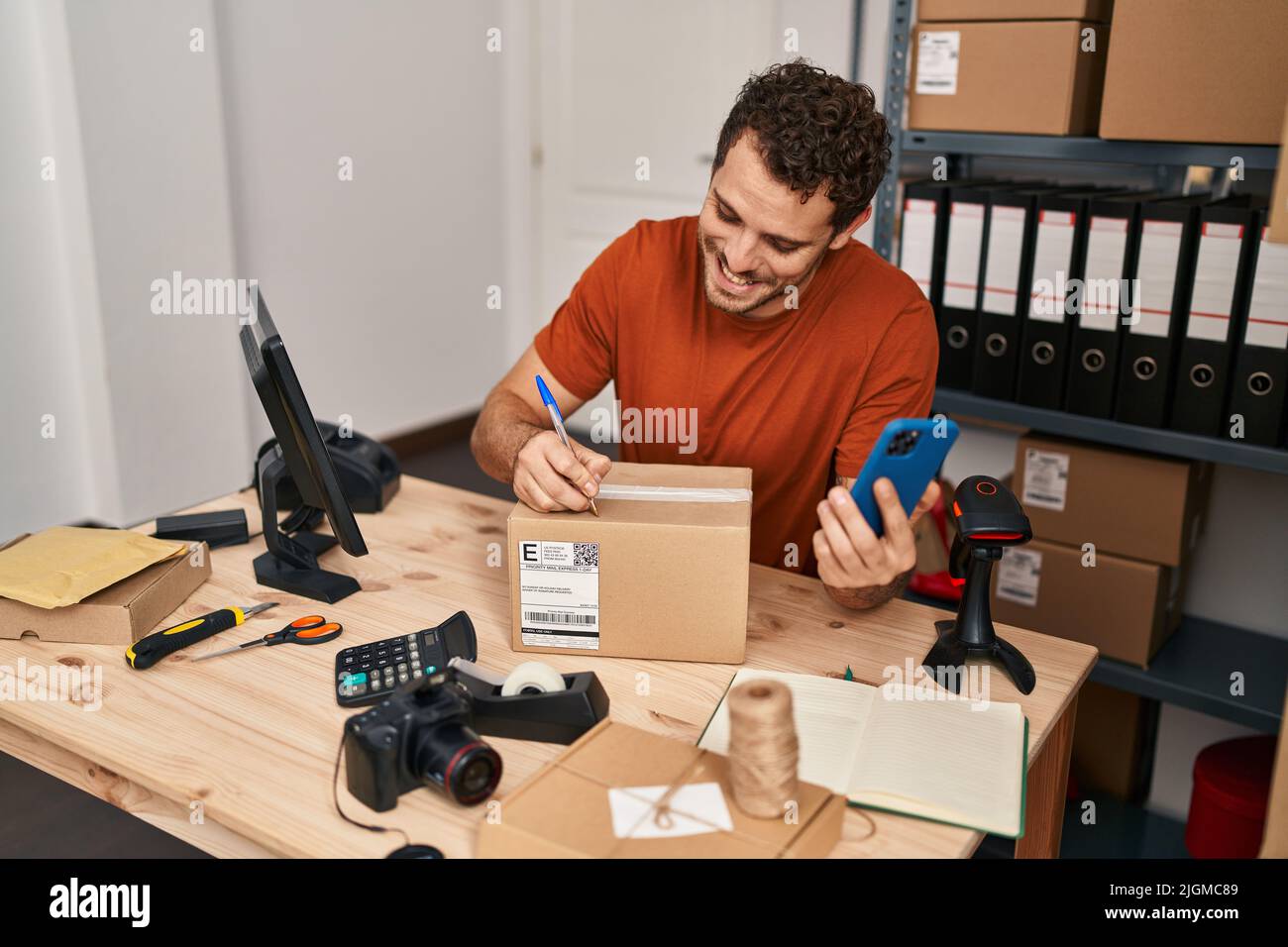 Young hispanic man ecommerce business worker write on package looking ...
