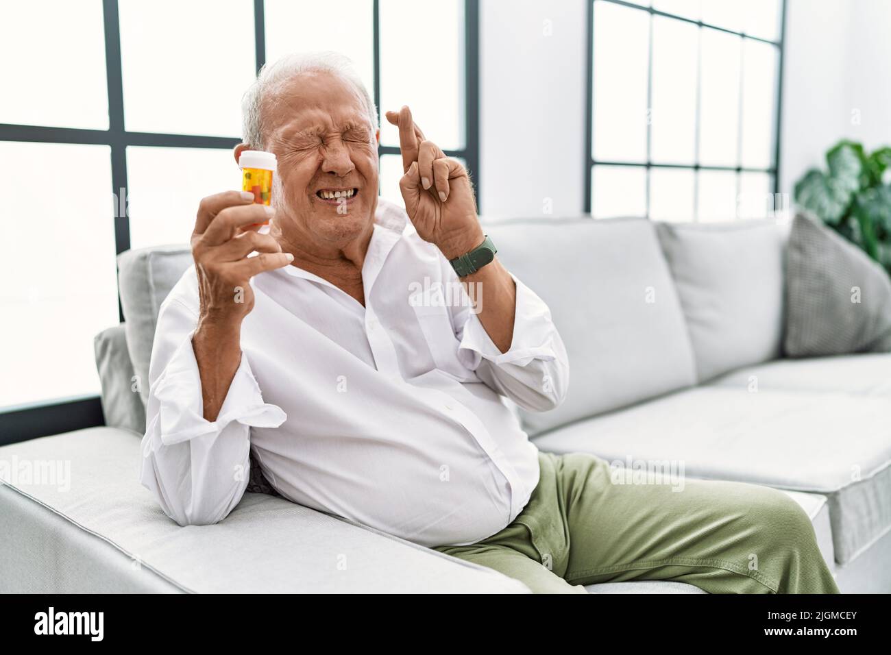 Senior man holding pills gesturing finger crossed smiling with hope and ...