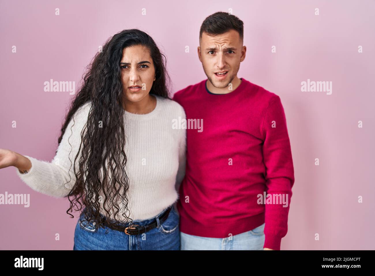 Young hispanic couple standing over pink background in shock face ...