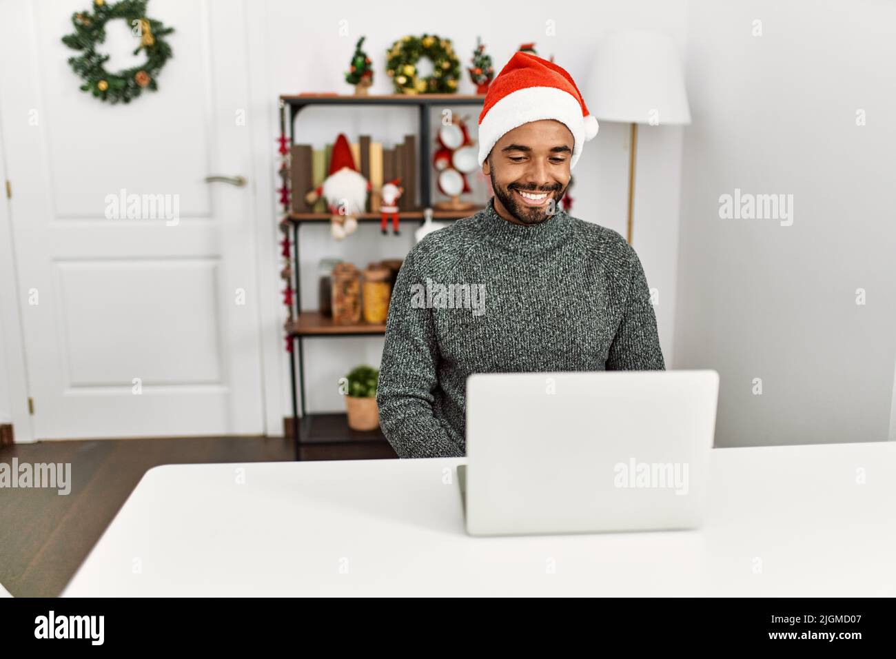 Young hispanic man with beard wearing christmas hat using laptop ...