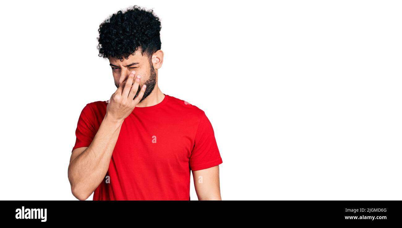 Young arab man with beard wearing casual red t shirt smelling something ...