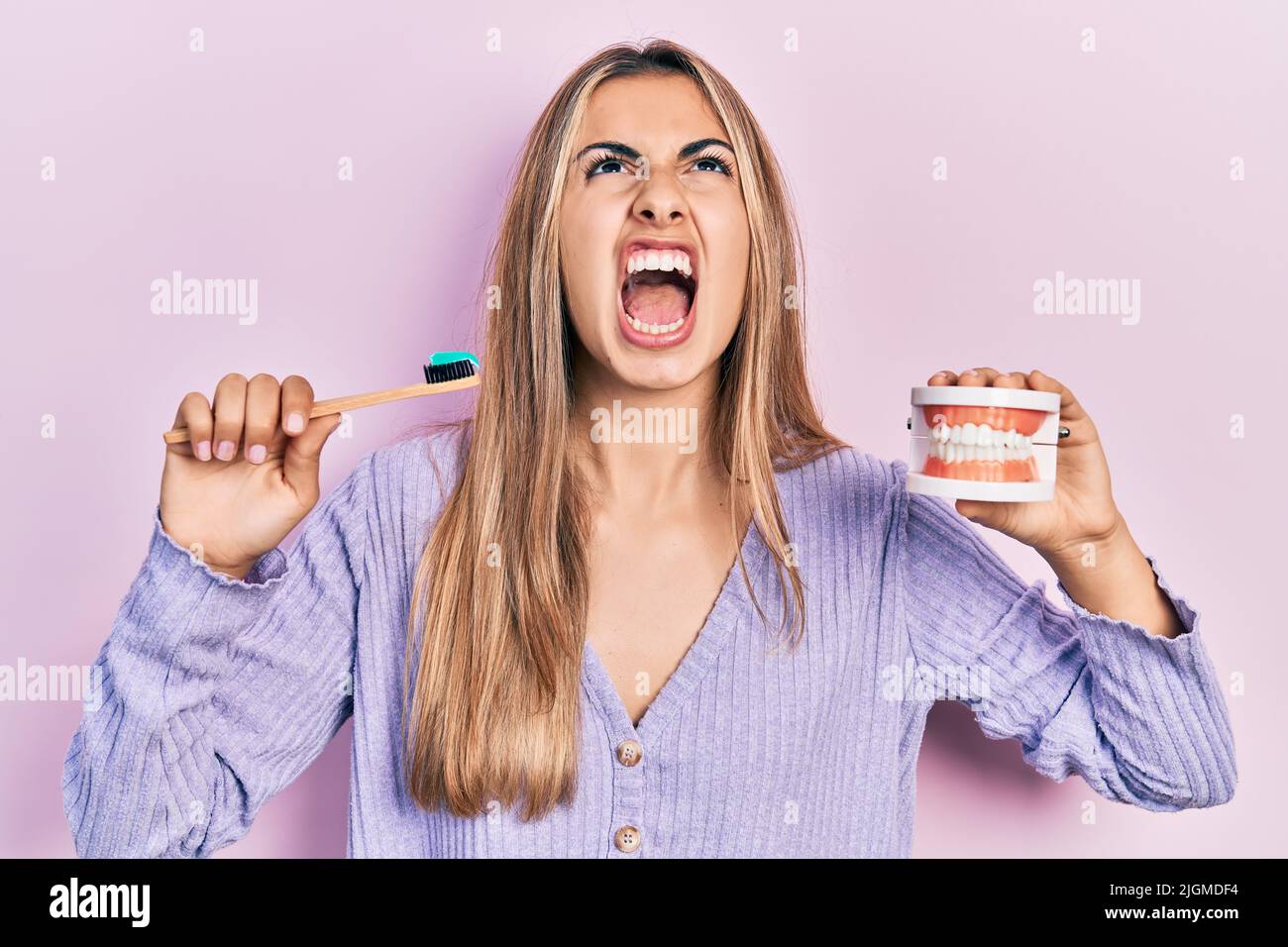 Beautiful hispanic woman holding denture and toothbrush angry and mad ...