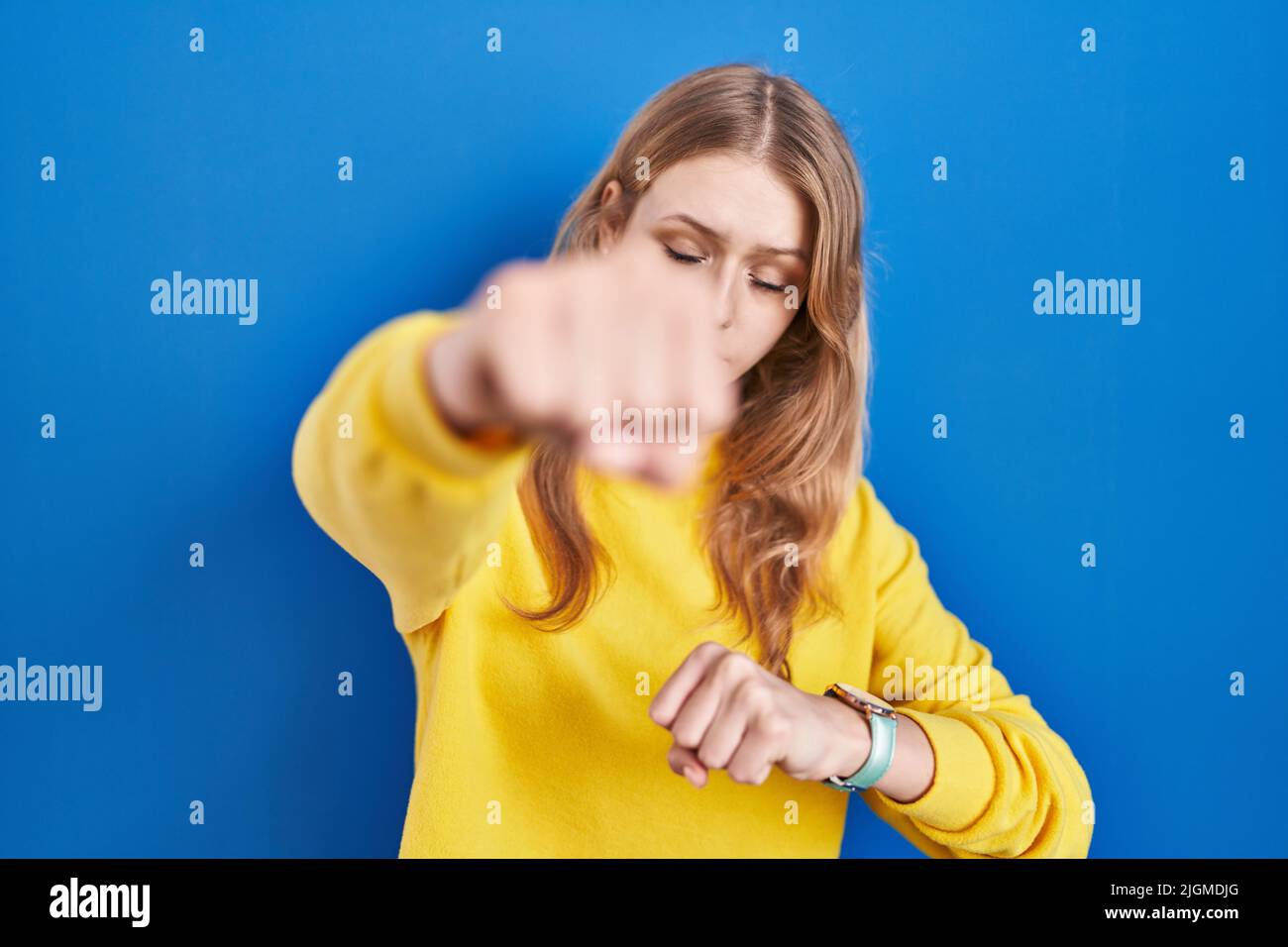 Young caucasian woman standing over blue background punching fist to ...