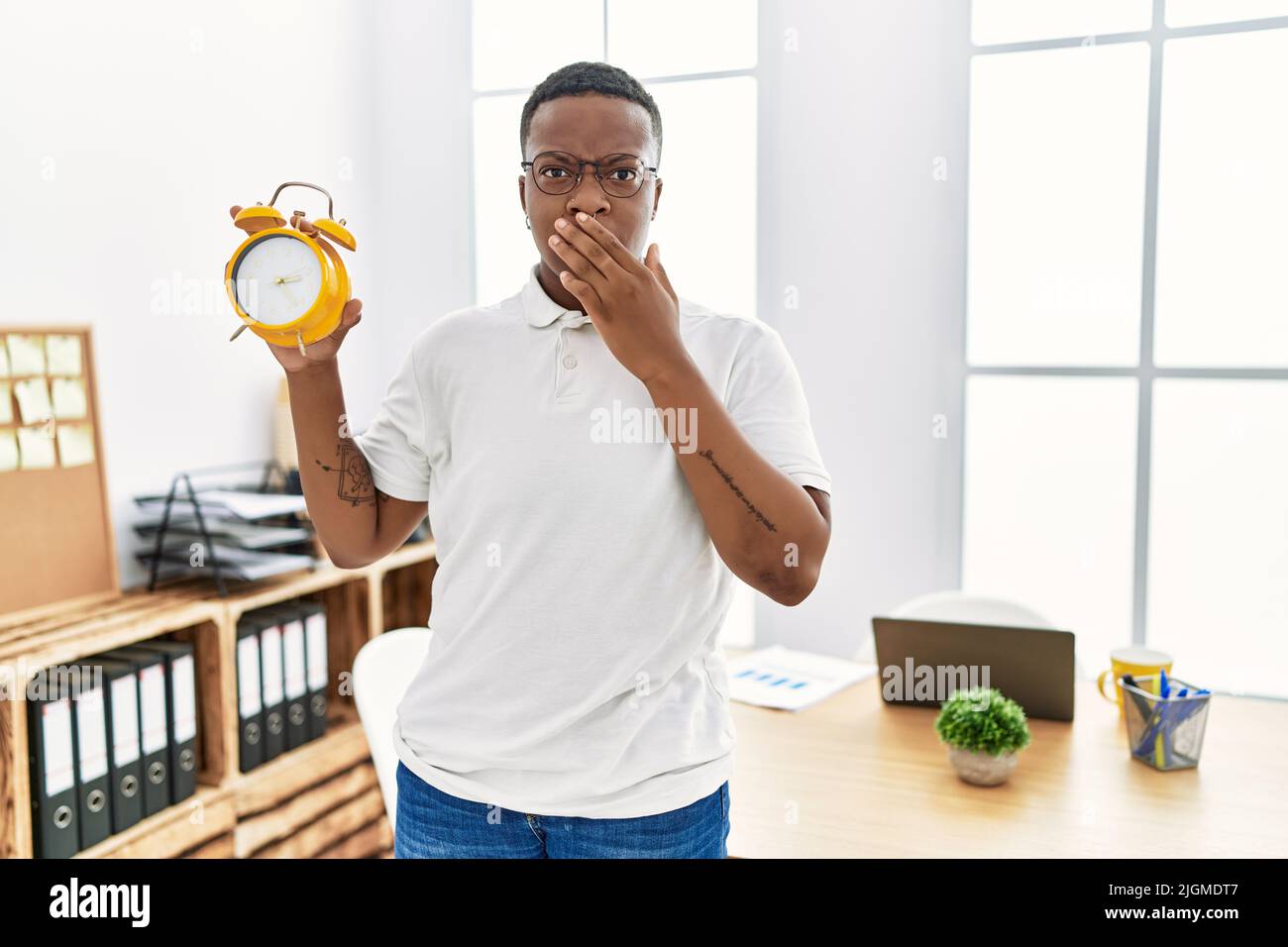 Young african man holding alarm clock at the office covering mouth with ...
