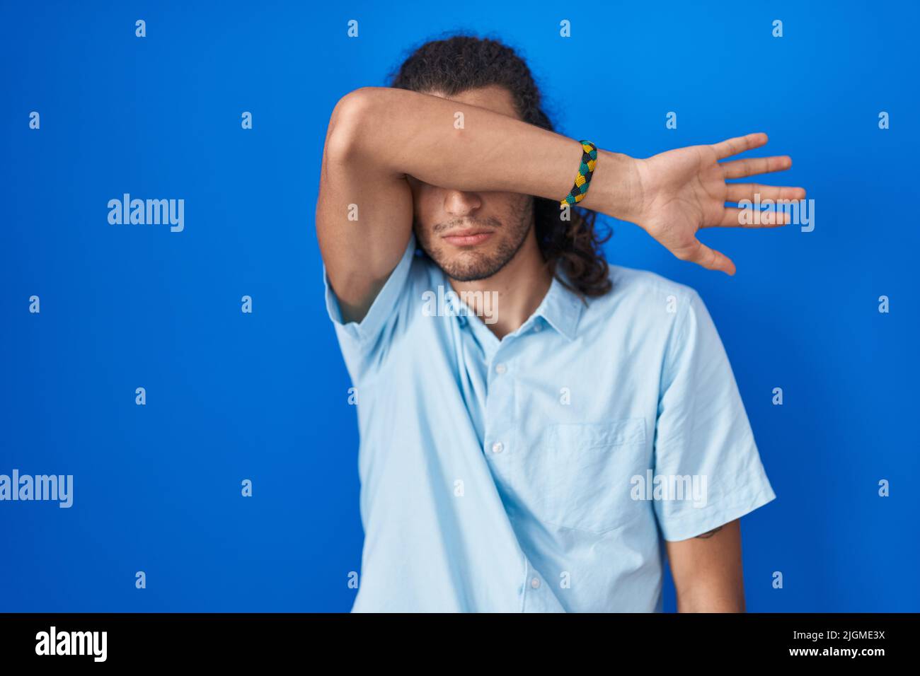 Young hispanic man standing over blue background covering eyes with arm ...