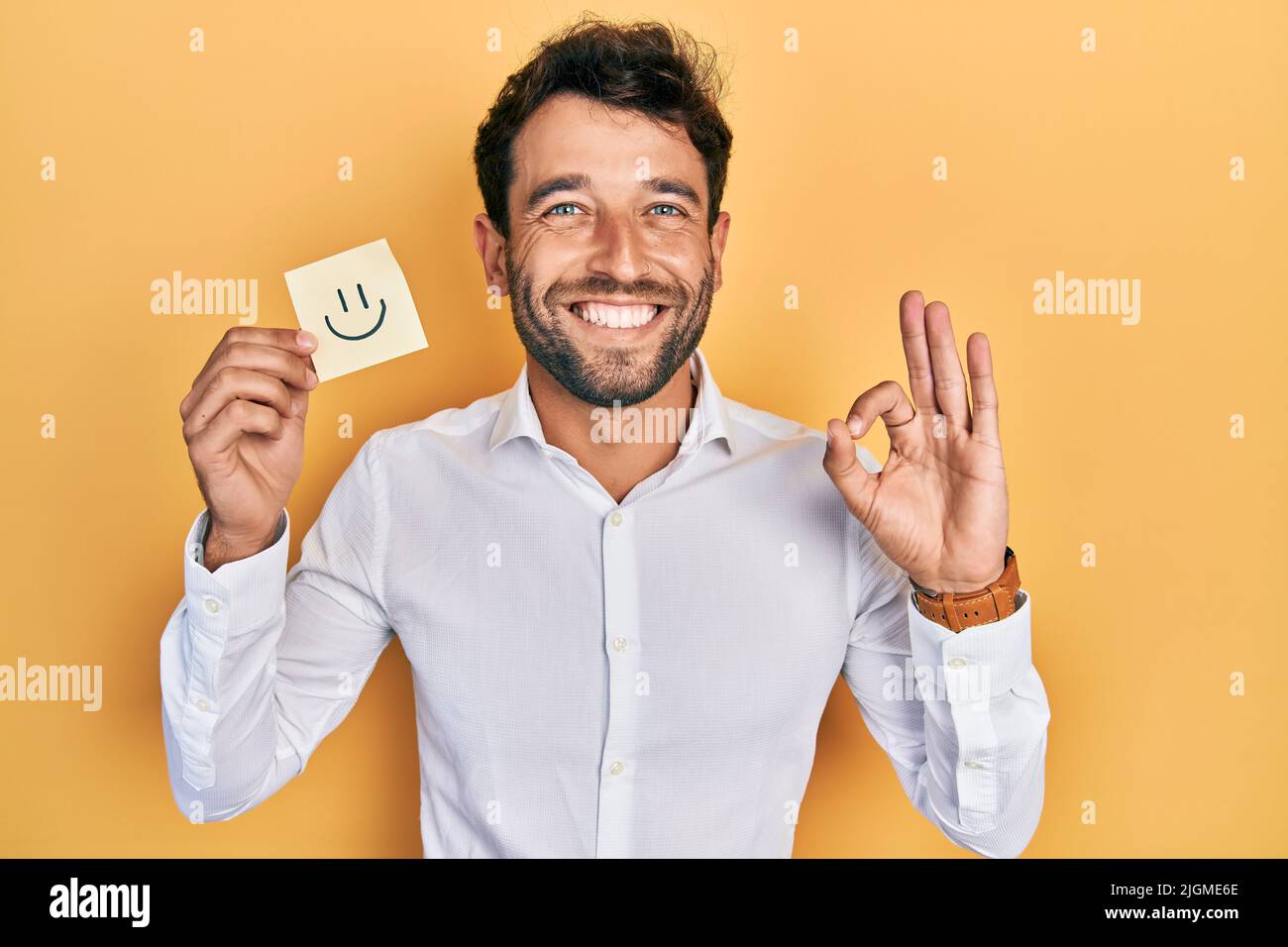 Handsome man with beard holding smile emoji reminder doing ok sign with ...