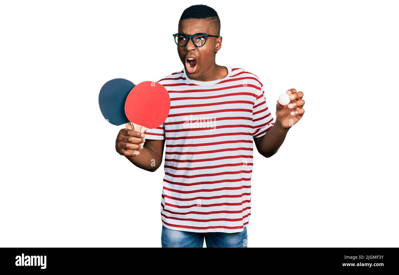Young african american man holding red ping pong rackets and ball angry ...
