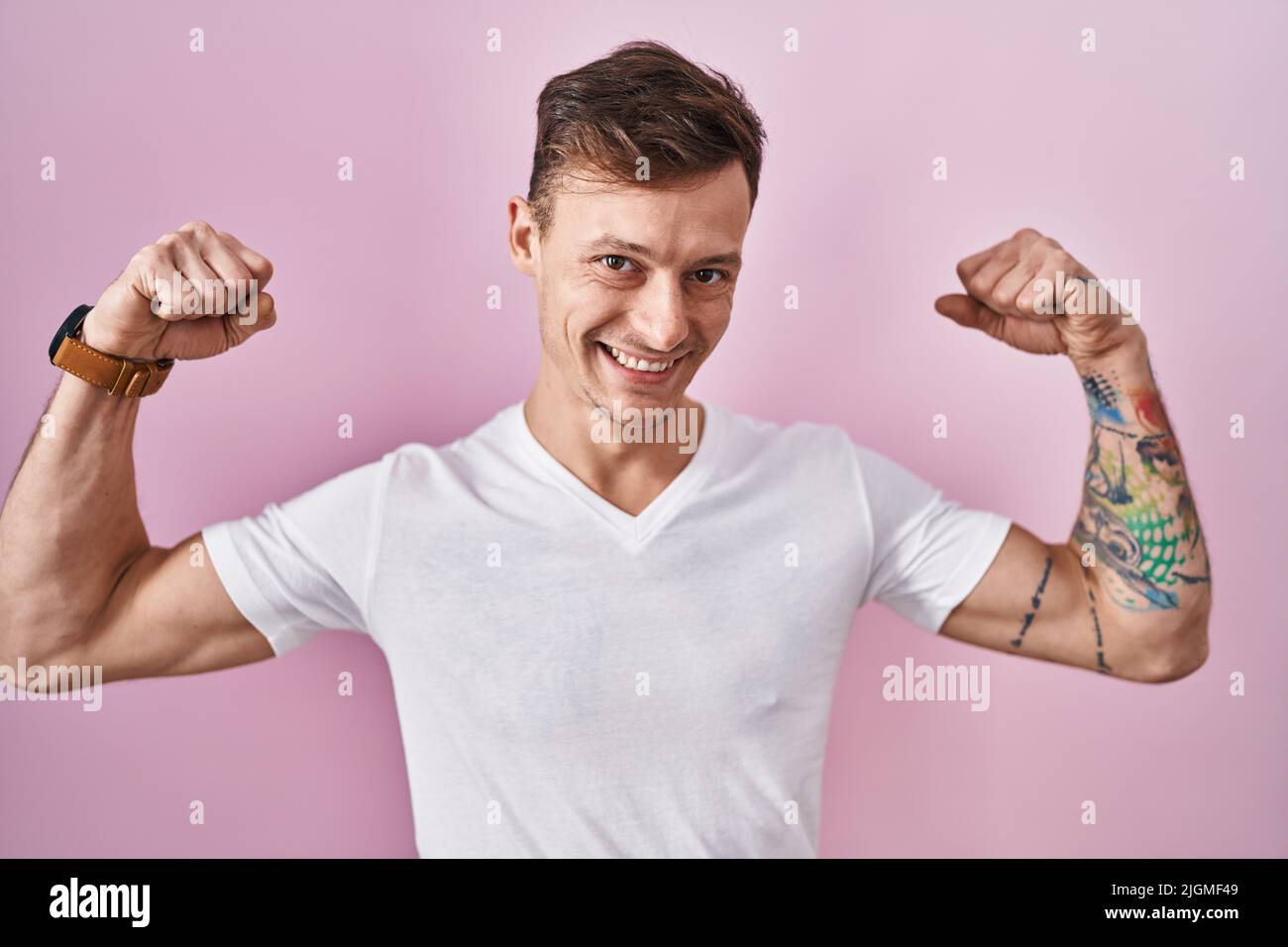 Caucasian man standing over pink background showing arms muscles ...