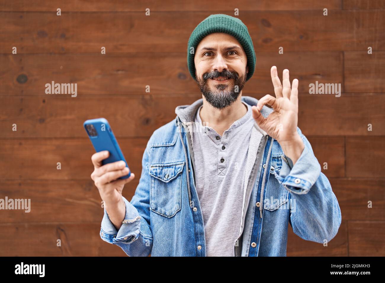 Young hispanic man using smartphone doing ok sign with fingers, smiling ...
