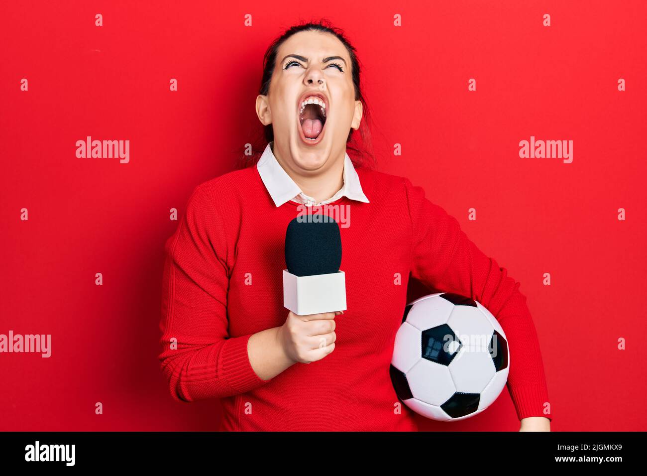Young hispanic woman holding reporter microphone and soccer ball angry ...