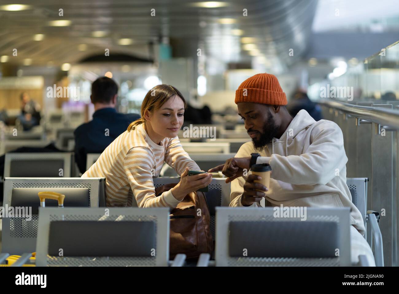 Young international couple using mobile phone while sitting in airport terminal waiting for ...