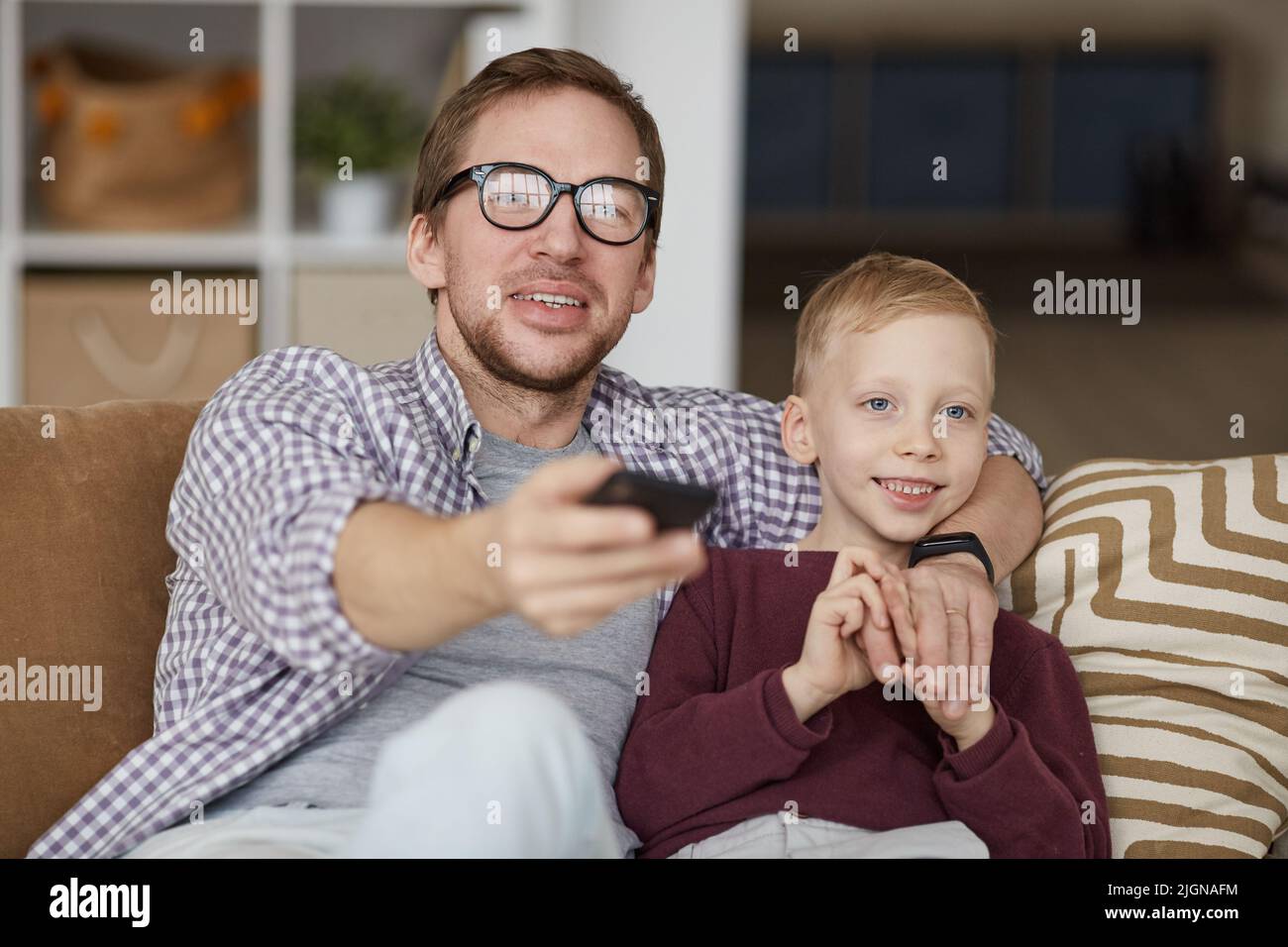 Smiling relaxed father embracing son around neck and changing channel ...
