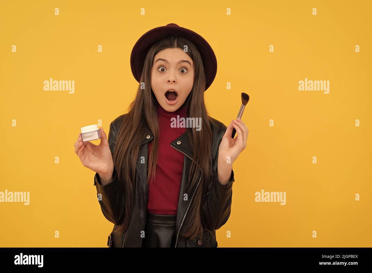 shocked kid in hat hold makeup brush and powder on yellow background ...