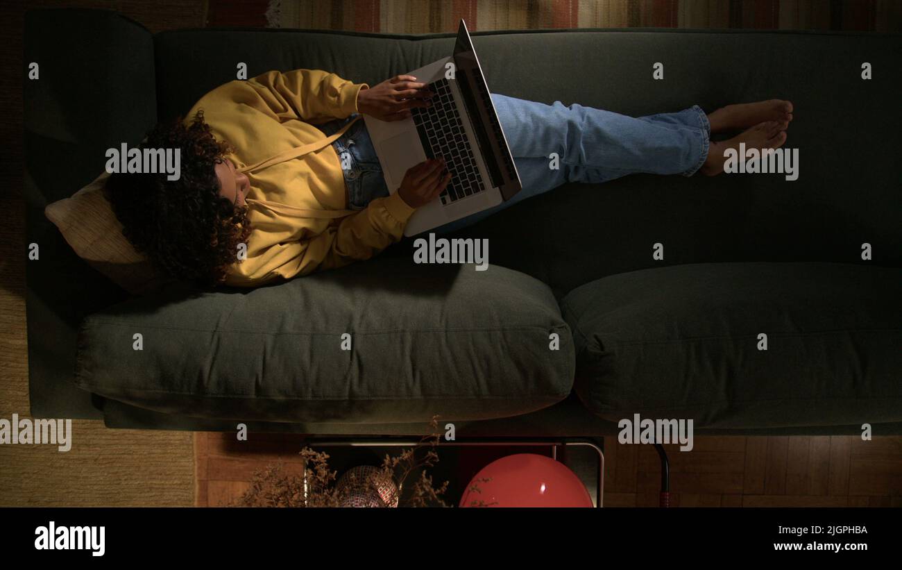 One young black girl lying down on couch opening laptop. Top view angle ...