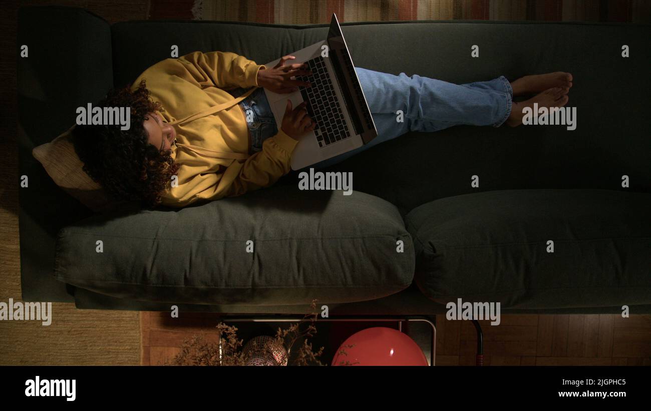 One young black girl lying down on couch opening laptop. Top view angle ...