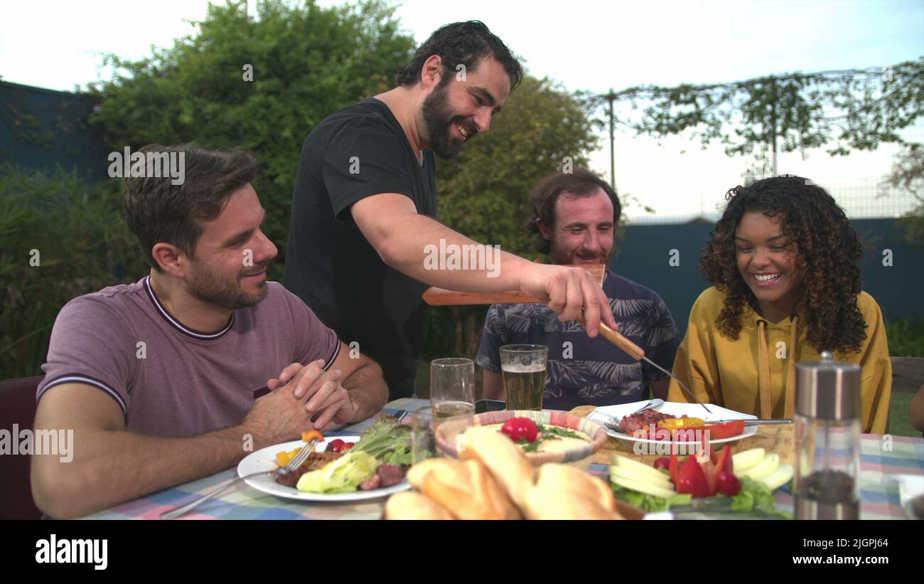 Group of cheerful friends eating barbecue together. BBQ chef serving food to friend. People ...