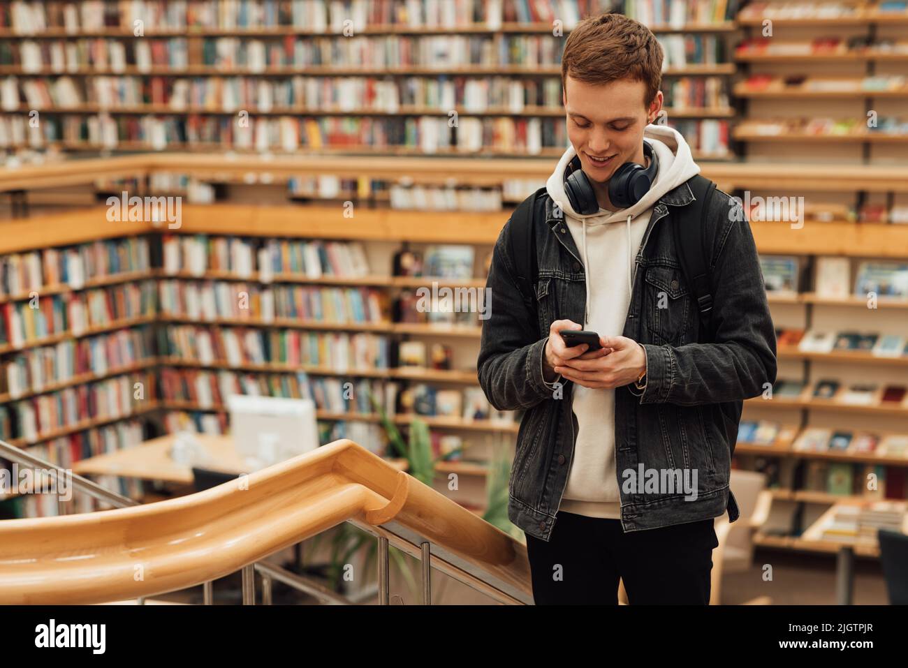 Smiling guy with a smartphone in a library. Student with headphones ...