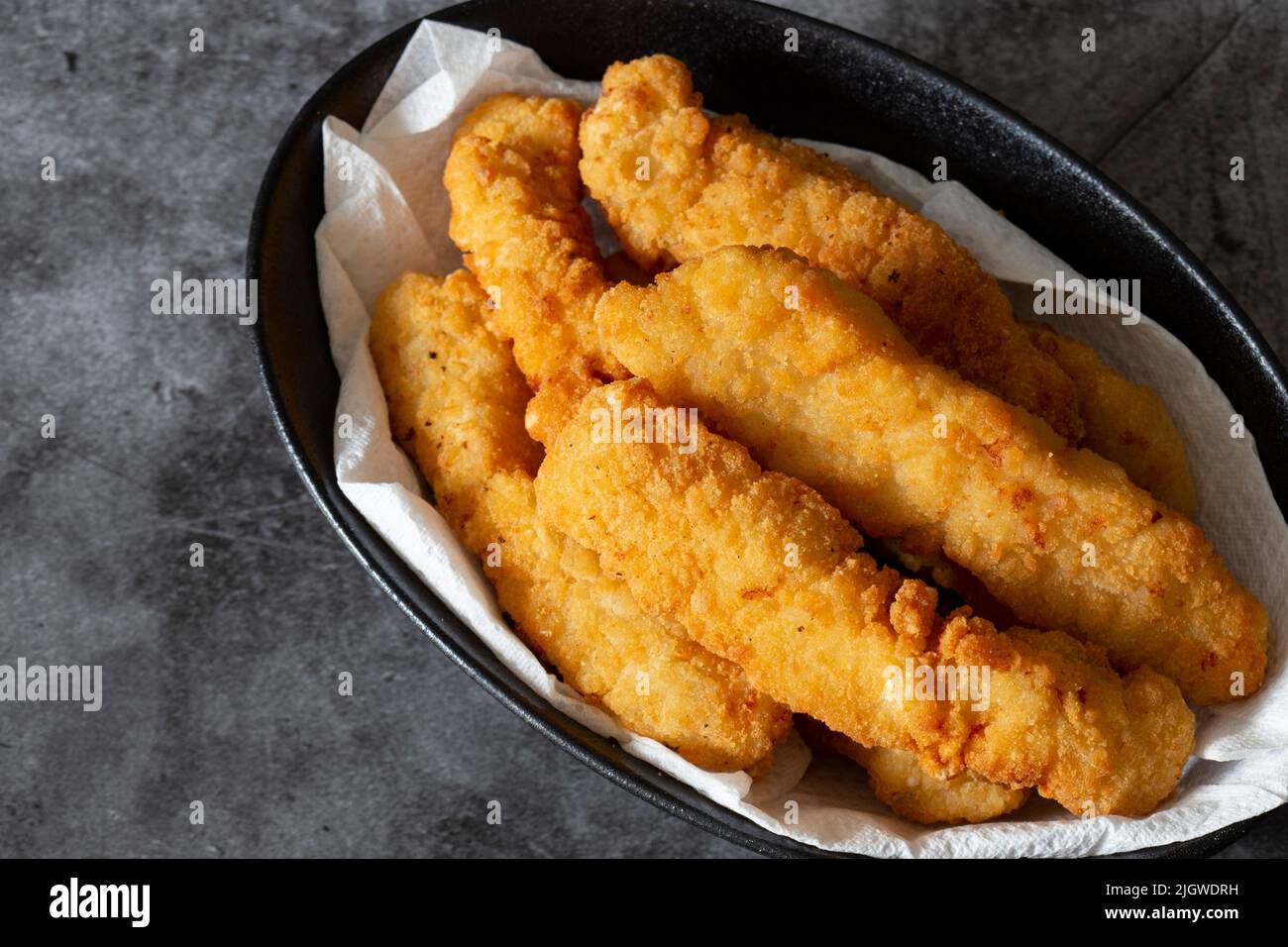 Breaded chicken fillet strips on a paper towel in a black bowl. On a ...