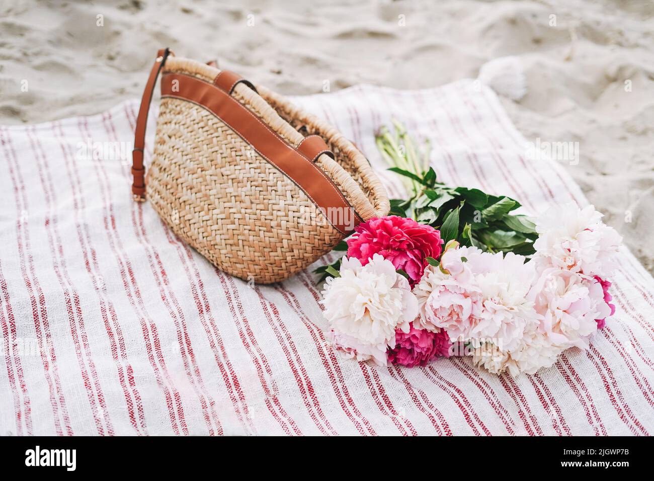 Wicker bag and bouquet of pink and white peonies on blanket on the sand ...