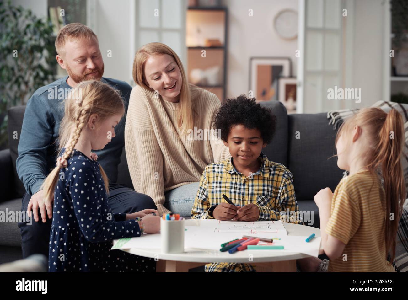 Adoptive children drawing together at table while parents admiring ...
