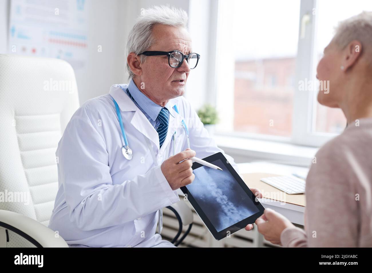 Confident senior male doctor in lab coat using digital tablet while ...