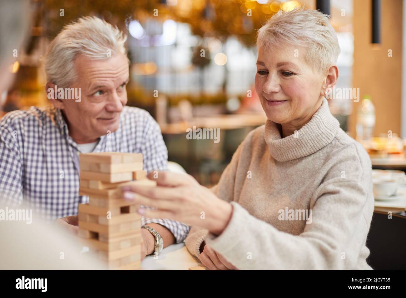 Smiling well-adjusted senior woman in warm sweater taking toy block out ...