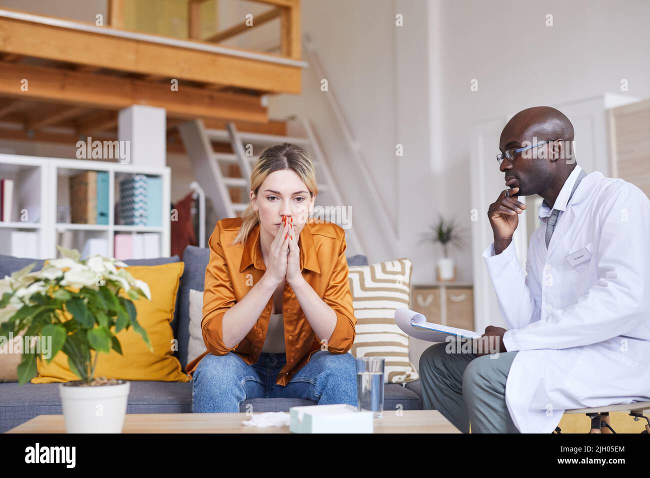 Pensive puzzled young woman in orange blouse sitting on sofa and ...