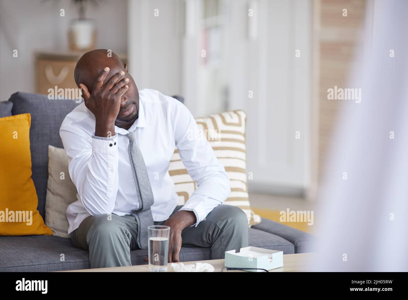 Confused young Black male patient in formalwear sitting on sofa and ...