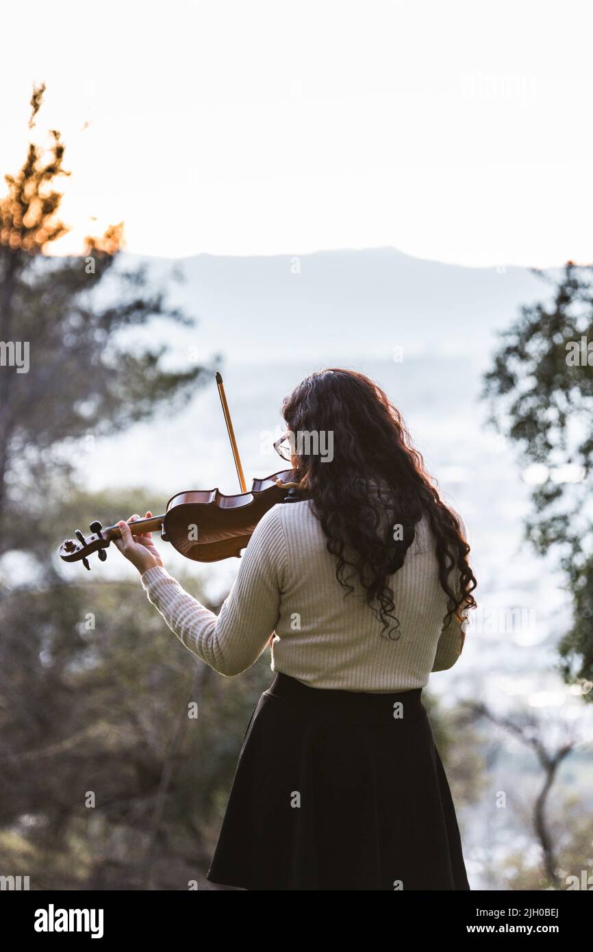 Brunette woman seen from back playing violin outside in the mountain ...