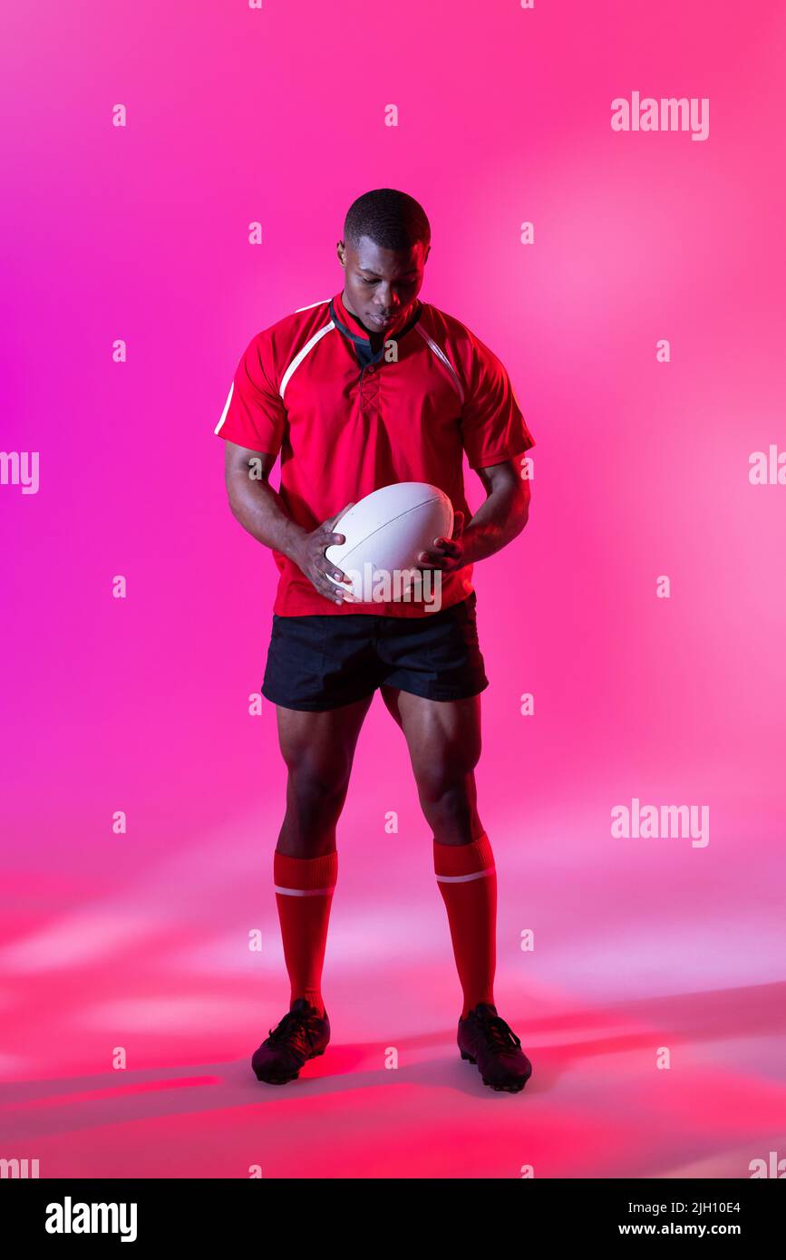 African american male rugby player with rugby ball over pink lighting ...